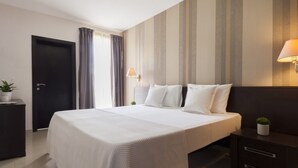 Quarto de hotel com uma cama grande, uma mesa de cabeceira com abajur, uma mesa de apoio escura e um vaso com planta.