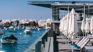 Varanda de hotel à beira-mar com espreguiçadeiras e guarda-sóis, com vista para uma marina e barcos.
