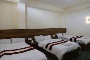 Quarto de hotel com três camas, cada uma com roupa de cama branca e detalhes vermelhos, cabeceira de madeira e mesinha de cabeceira com luminária.