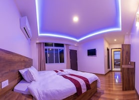 Quarto moderno com cabeceira de madeira, cama grande com roupa de cama branca e detalhes vermelhos, janela com cortinas e corredor que dá para outro cômodo.