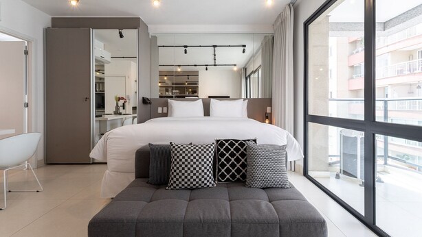 Quarto moderno com uma cama grande, um banco estofado cinza e vista da cidade através de amplas janelas.