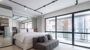 Quarto moderno com uma cama grande, um banquinho cinzento e vista da cidade através de grandes janelas.