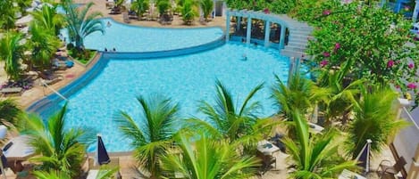 Uma piscina grande e convidativa cercada por palmeiras e espreguiçadeiras, com vista para edifícios de hotel ao fundo.