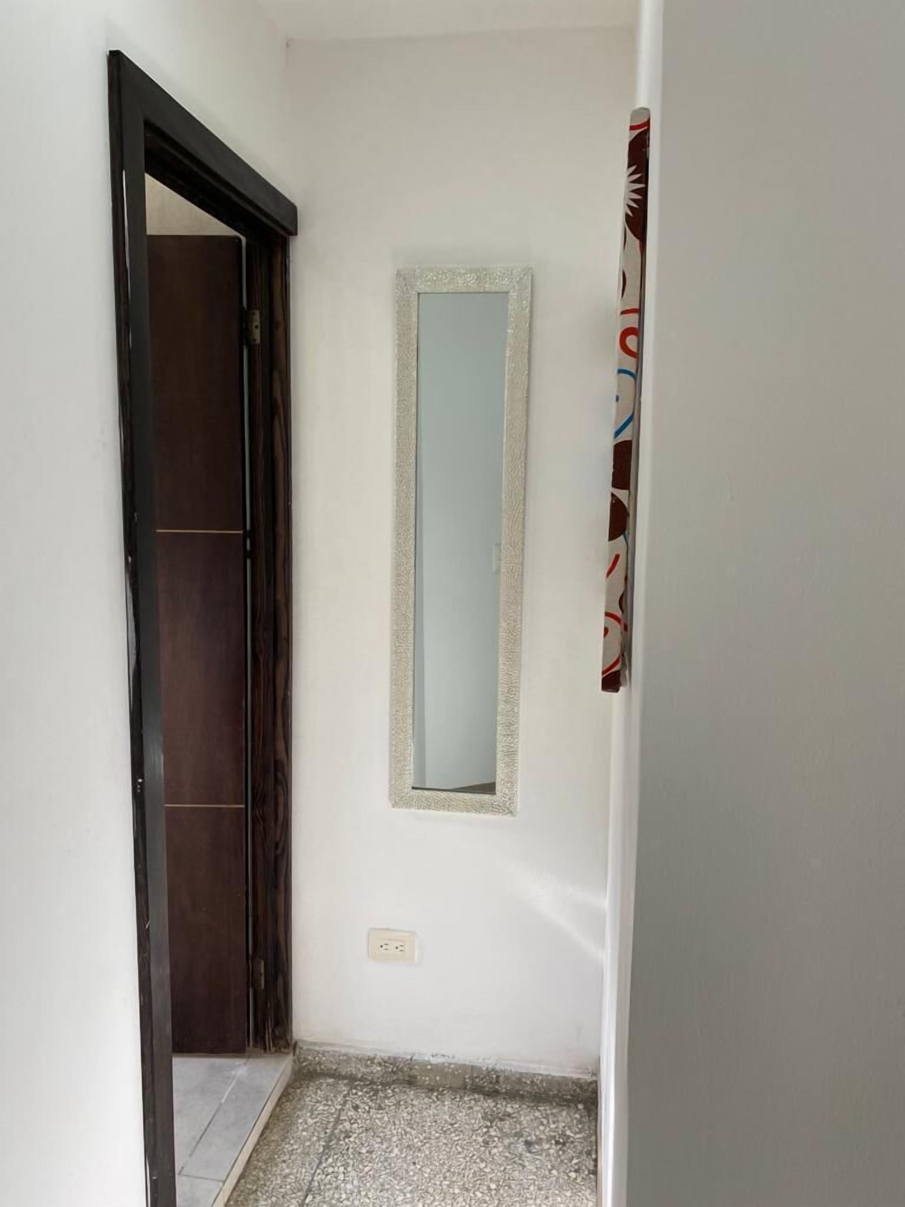 Um corredor estreito com uma porta de madeira à esquerda, um pequeno espelho na parede e uma cortina estampada à direita.