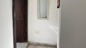 Um corredor estreito com uma porta de madeira à esquerda, um pequeno espelho na parede e uma cortina estampada à direita.