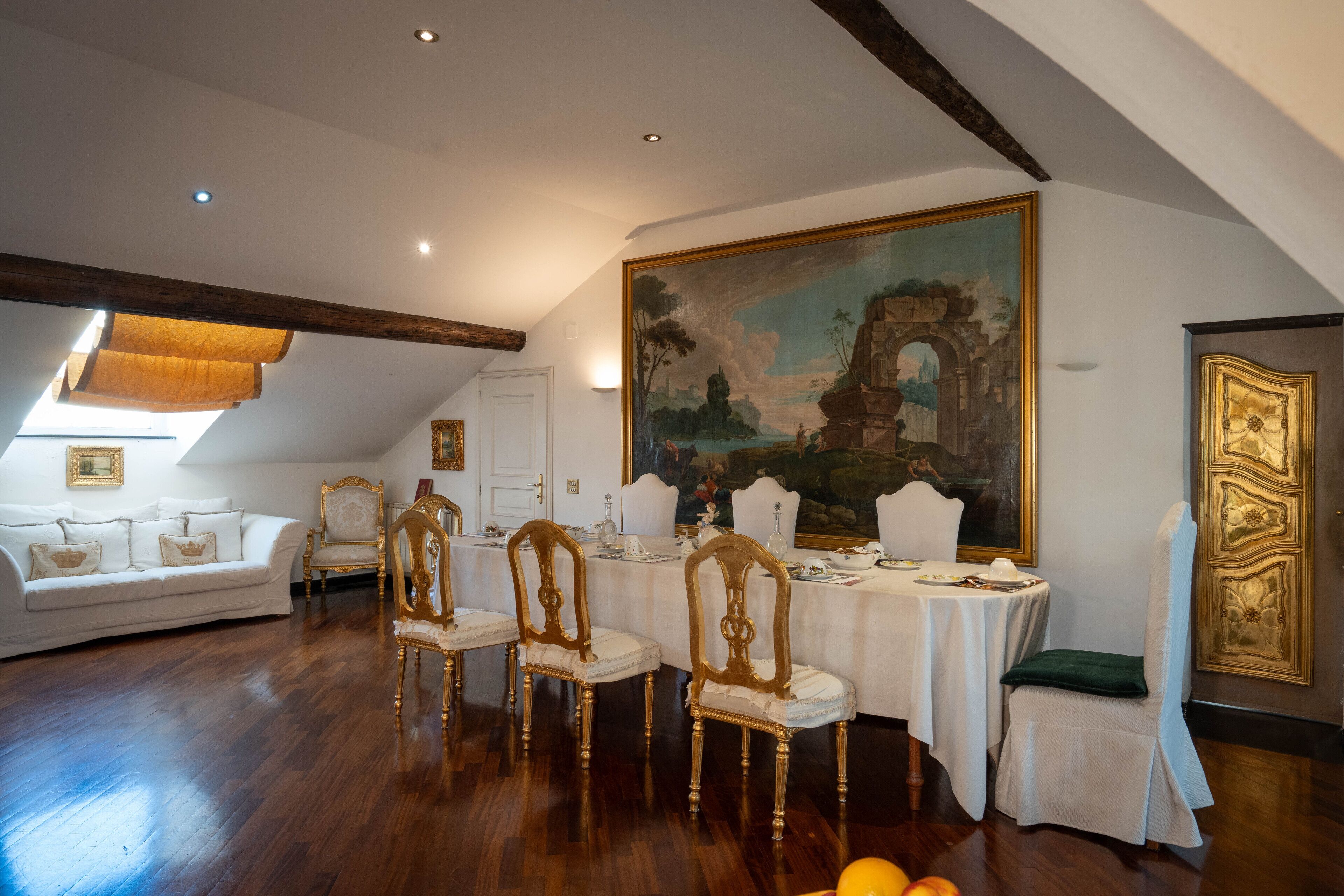 Uma sala de jantar com uma mesa posta para quatro pessoas, rodeada por cadeiras elaboradas, um quadro grande na parede e uma vista do exterior através de um clarabóia.