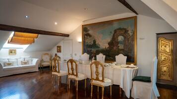 Uma sala de jantar com uma mesa posta para quatro pessoas, rodeada por cadeiras elaboradas, um quadro grande na parede e uma vista do exterior através de um clarabóia.