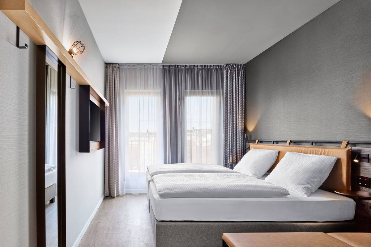 Quarto de hotel moderno com uma cama grande, cabeceira de madeira, televisão de tela plana na parede e uma janela com cortinas leves.