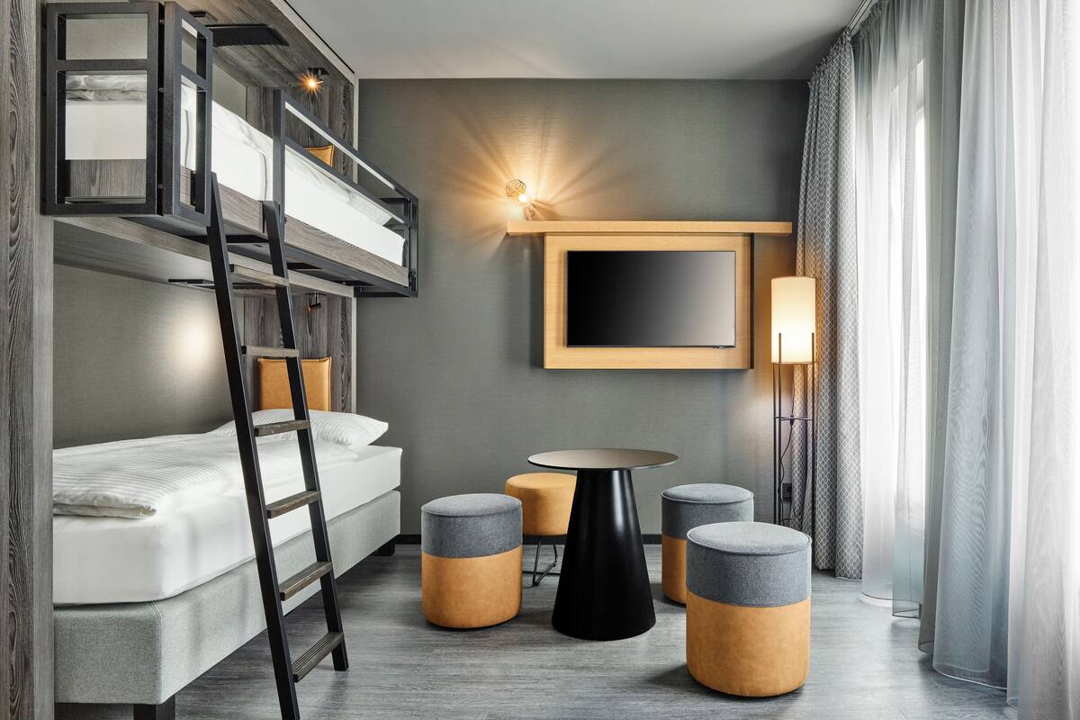 Quarto de hotel moderno com beliche, televisão de tela plana na parede, mesa redonda e luminária de chão.