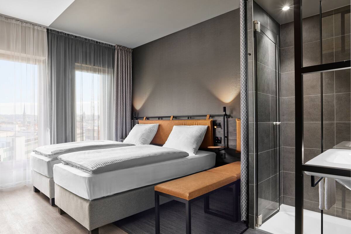 Quarto de hotel moderno com cama grande, um banco, mesa de cabeceira, área de chuveiro e vista para a cidade.