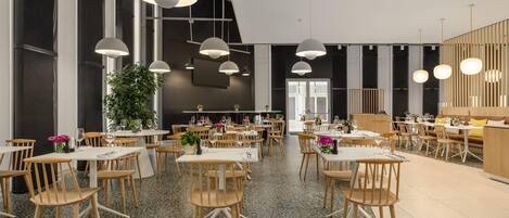 Uma sala de jantar ampla, com várias mesas e cadeiras, iluminação pendente e um grande arranjo de plantas.
