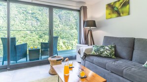 Uma sala de estar moderna com um sofá cinzento, uma mesa de centro de madeira e duas cadeiras. Há uma grande janela que oferece vista para uma área arborizada.