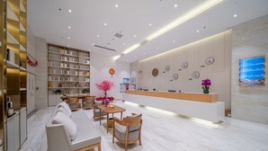 Um hall de hotel moderno com uma mesa de recepção, uma área de estar com sofá e poltronas, uma estante de livros e uma planta decorativa.