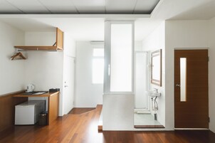 Quarto moderno e minimalista com piso de madeira, pequena cozinha compacta, armário de madeira, radiador branco e porta que dá para outro cômodo.
