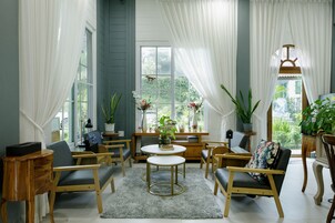 Um quarto aconchegante com uma mesa redonda, cadeiras de madeira e um sofá. Há janelas amplas com cortinas, plantas em vasos e vista para o verde lá fora.