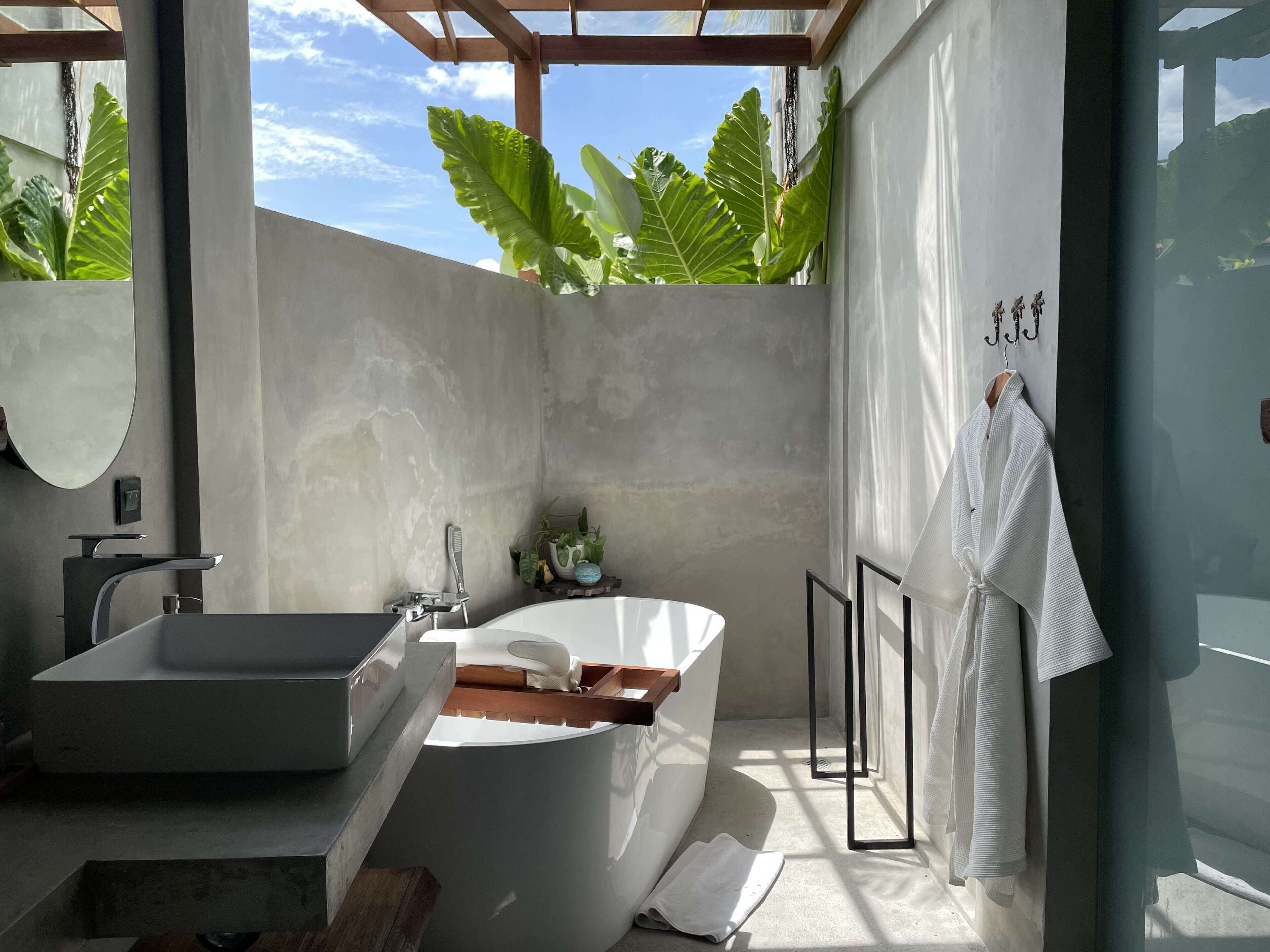 Um banheiro moderno com uma banheira livre, um espelho grande e uma pia. Há plantas na parede e um porta-toalhas com roupão.