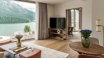 Uma sala de estar moderna com uma grande janela com vista para um lago e montanhas, uma televisão de tela plana e um espaço de refeições com uma mesa e cadeiras de madeira.