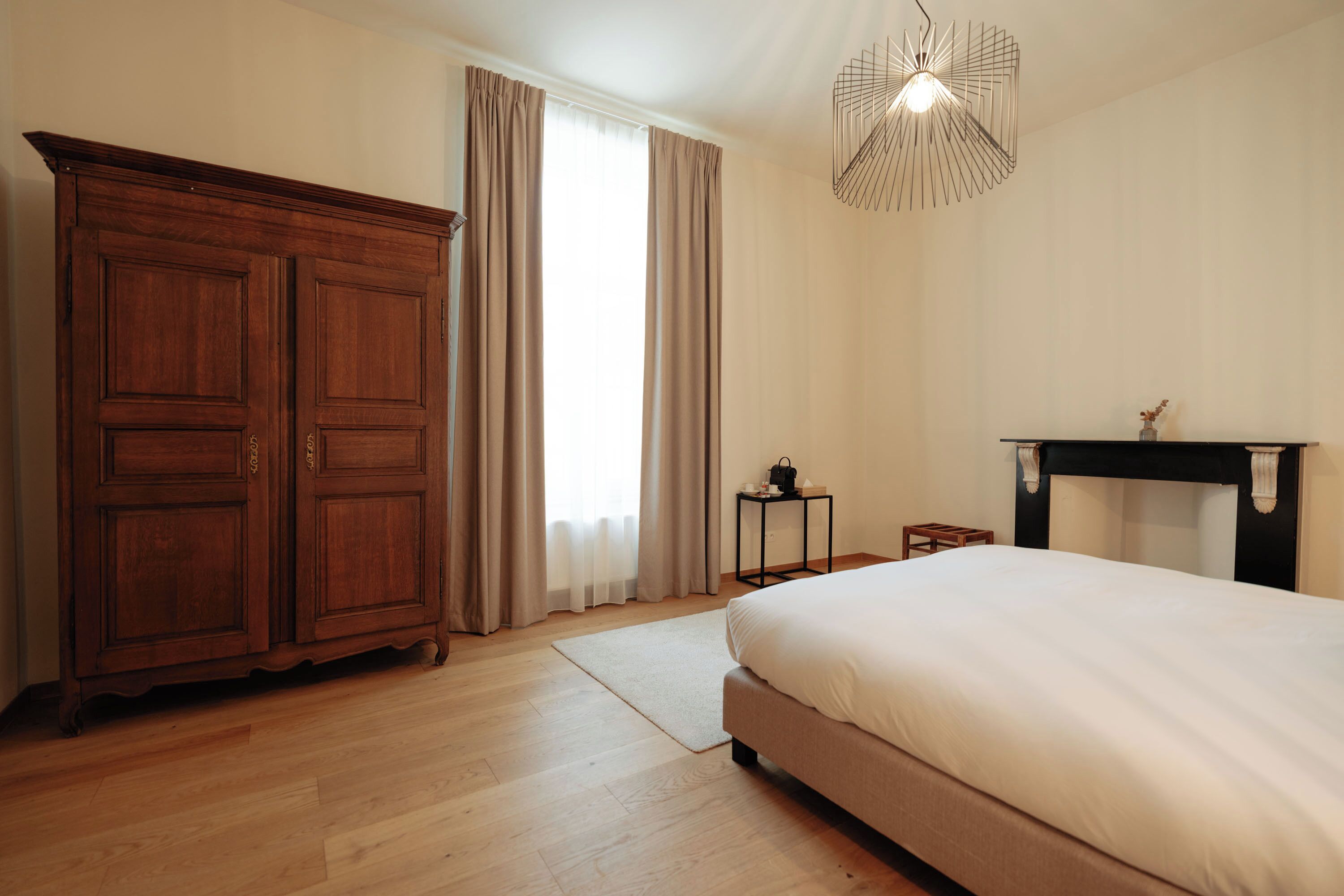 Quarto com um guarda-roupa de madeira grande, cama com roupa de cama branca, mesinha de cabeceira pequena, abajur e manto de lareira.