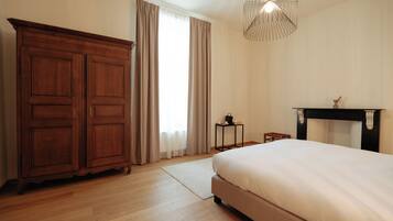 Quarto com um guarda-roupa de madeira grande, cama com roupa de cama branca, mesinha de cabeceira pequena, abajur e manto de lareira.