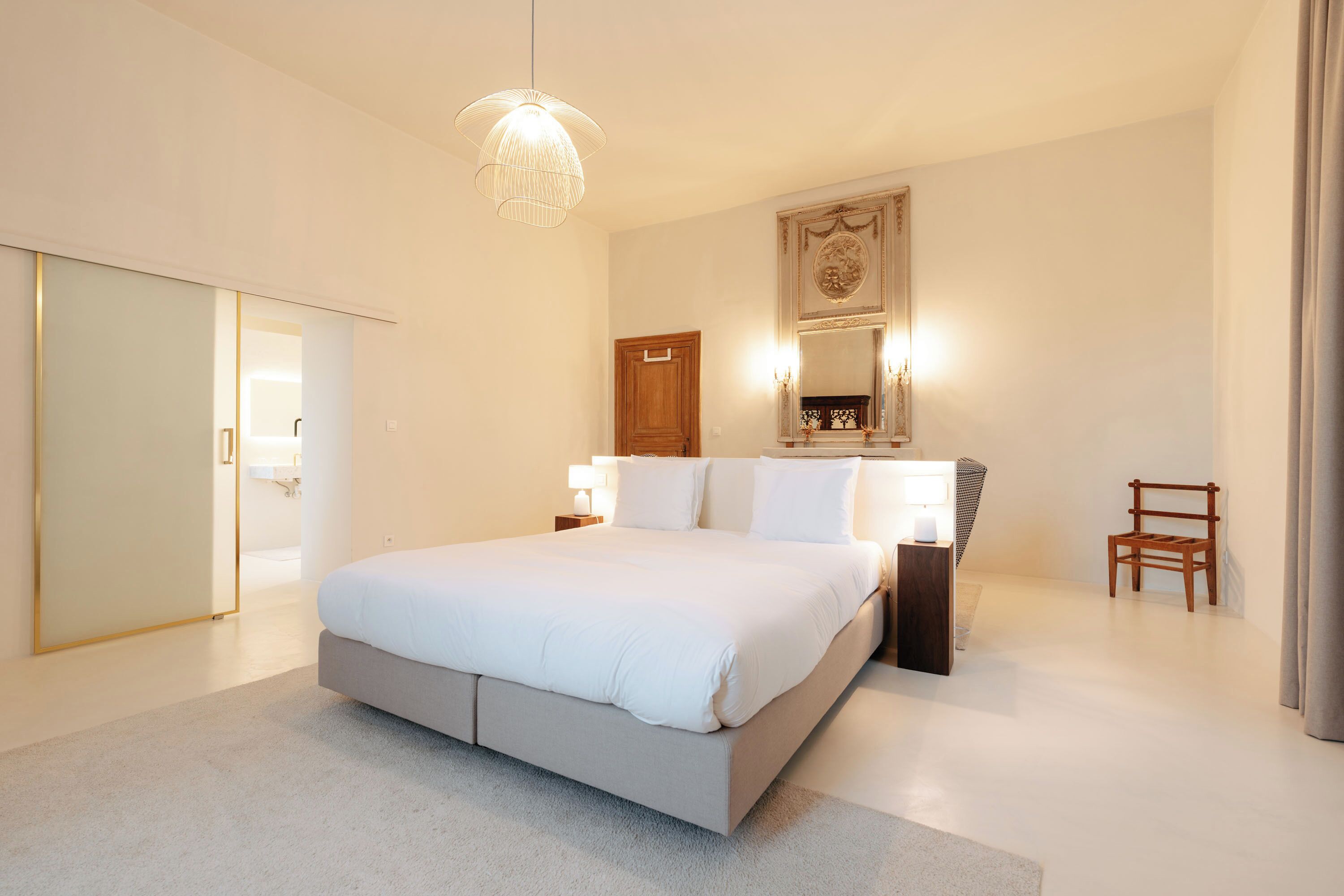 Quarto moderno com cama grande, luminárias de cabeceira, uma cadeira de madeira, espelho e porta de correr.