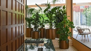 Um corredor com design geométrico no teto, plantas em vasos, uma mesa de vidro e vista para o exterior.