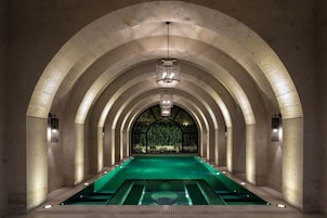 Um longo corredor arqueado, com piscina central, colunas de pedra e um candelabro.