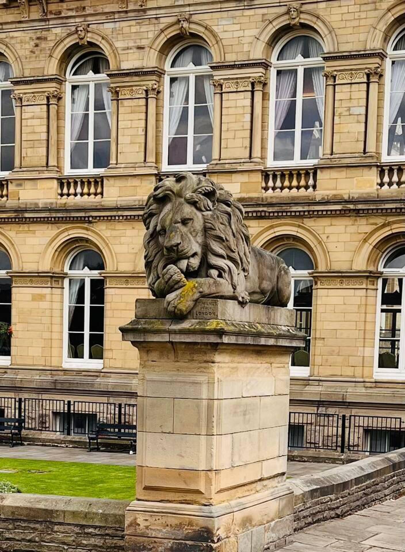 Estátua de leão de pedra em um pedestal, em frente a um edifício com janelas em arco e fachada de tijolos.