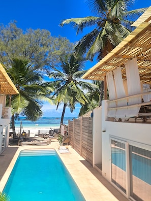 Área de piscina com vista para a praia e oceano, cercada por palmeiras e um muro de madeira.