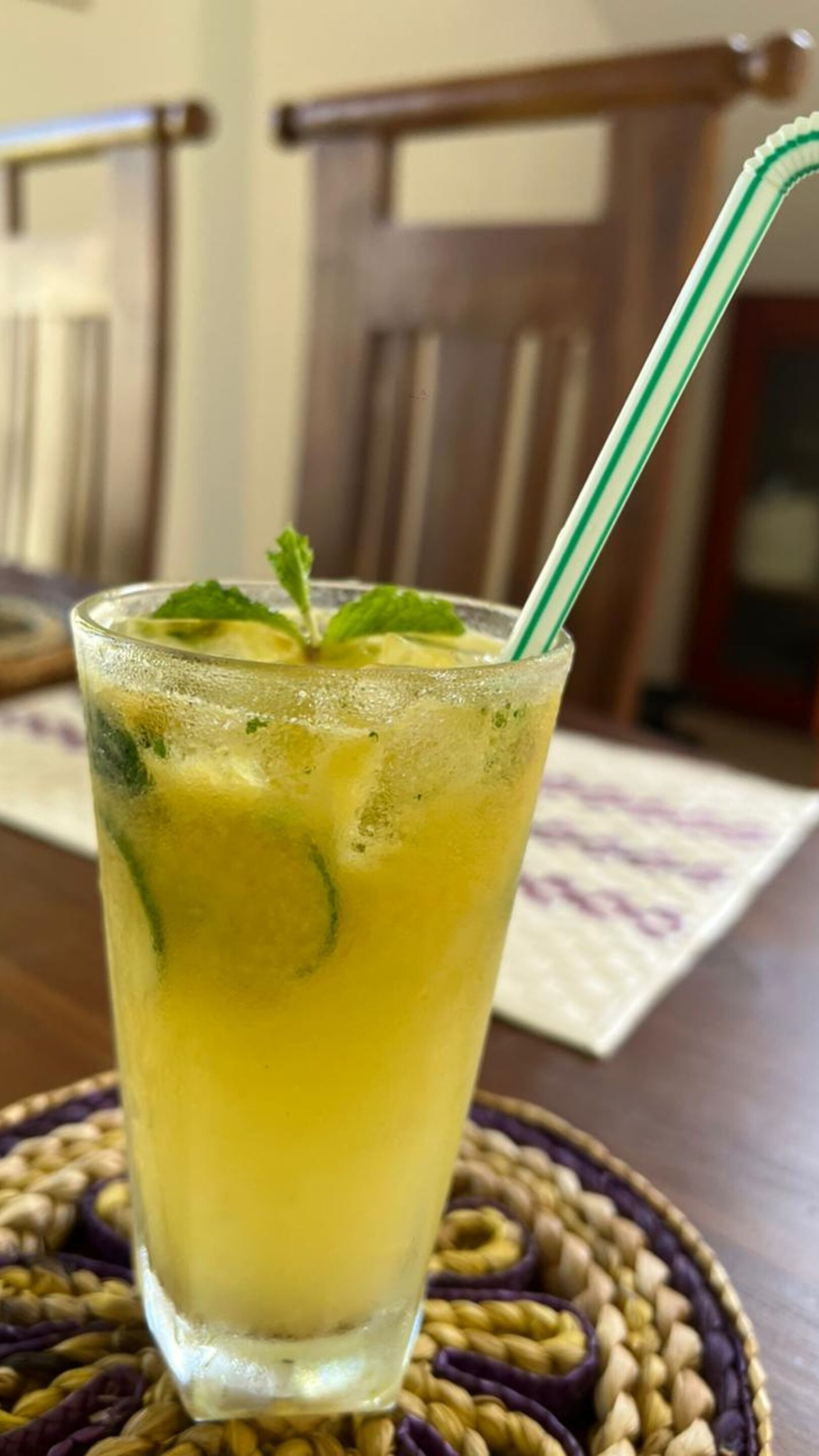 Um copo de bebida amarela com folhas de hortelã e um canudo listrado em verde e branco sobre um jogo americano trançado.