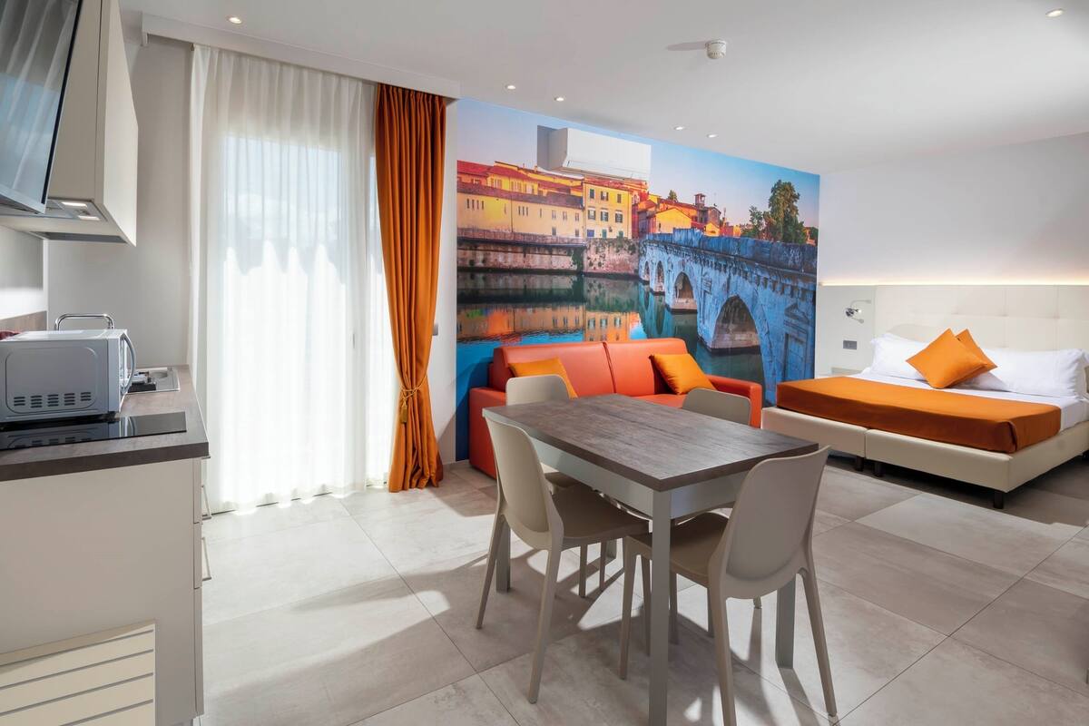 Quarto de hotel moderno com área de refeições, cama com roupa de cama laranja e um mural de uma ponte e edifícios.