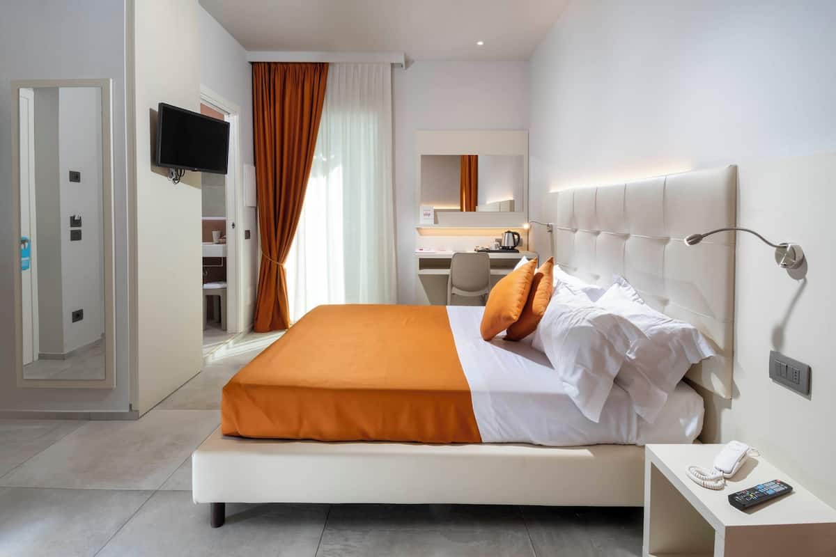 Quarto de hotel moderno com uma cama grande, uma mesinha de cabeceira com telefone e controle remoto, uma TV fixada na parede e um banheiro visível através de uma porta aberta.