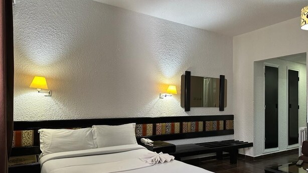 Quarto de hotel com uma cama grande, duas luminárias fixadas na parede, um espelho e um banco.