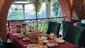 Mesa de café da manhã posta com pratos, xícaras e diversas opções de comida, rodeada por cadeiras verdes e uma toalha de mesa vermelha, com vista para o verde através de um arco.