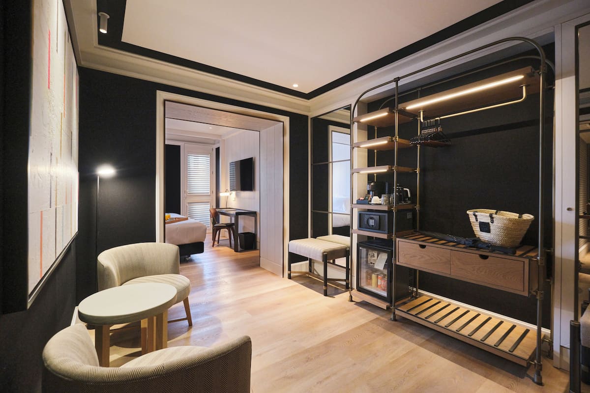 Quarto de hotel moderno com piso de madeira, área de estar com mesa e cadeiras, um banco embutido, guarda-roupa com prateleiras e um cabideiro com cestas.