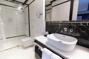 Casa de banho moderna com chuveiro envidraçado, lavatório branco e painel de fundo em mármore preto.