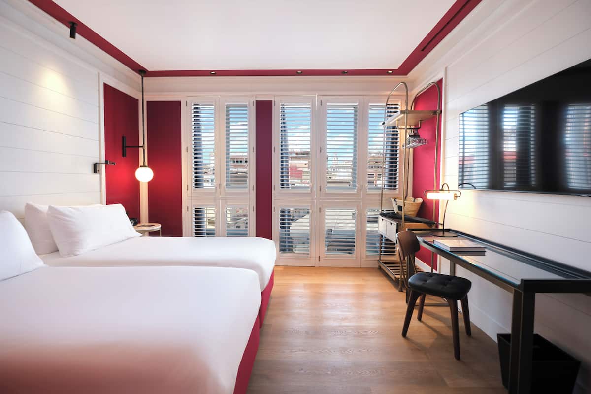 Quarto de hotel com duas camas, uma escrivaninha, uma cadeira, uma luminária e uma janela grande com persianas.