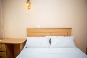 Cama bem arrumada com duas almofadas, mesa de cabeceira de madeira com gavetas e luminária fixada na parede.