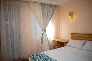 Quarto de hotel com cama, mesa de cabeceira, luminária fixada na parede e cortinas translúcidas com laço decorativo.