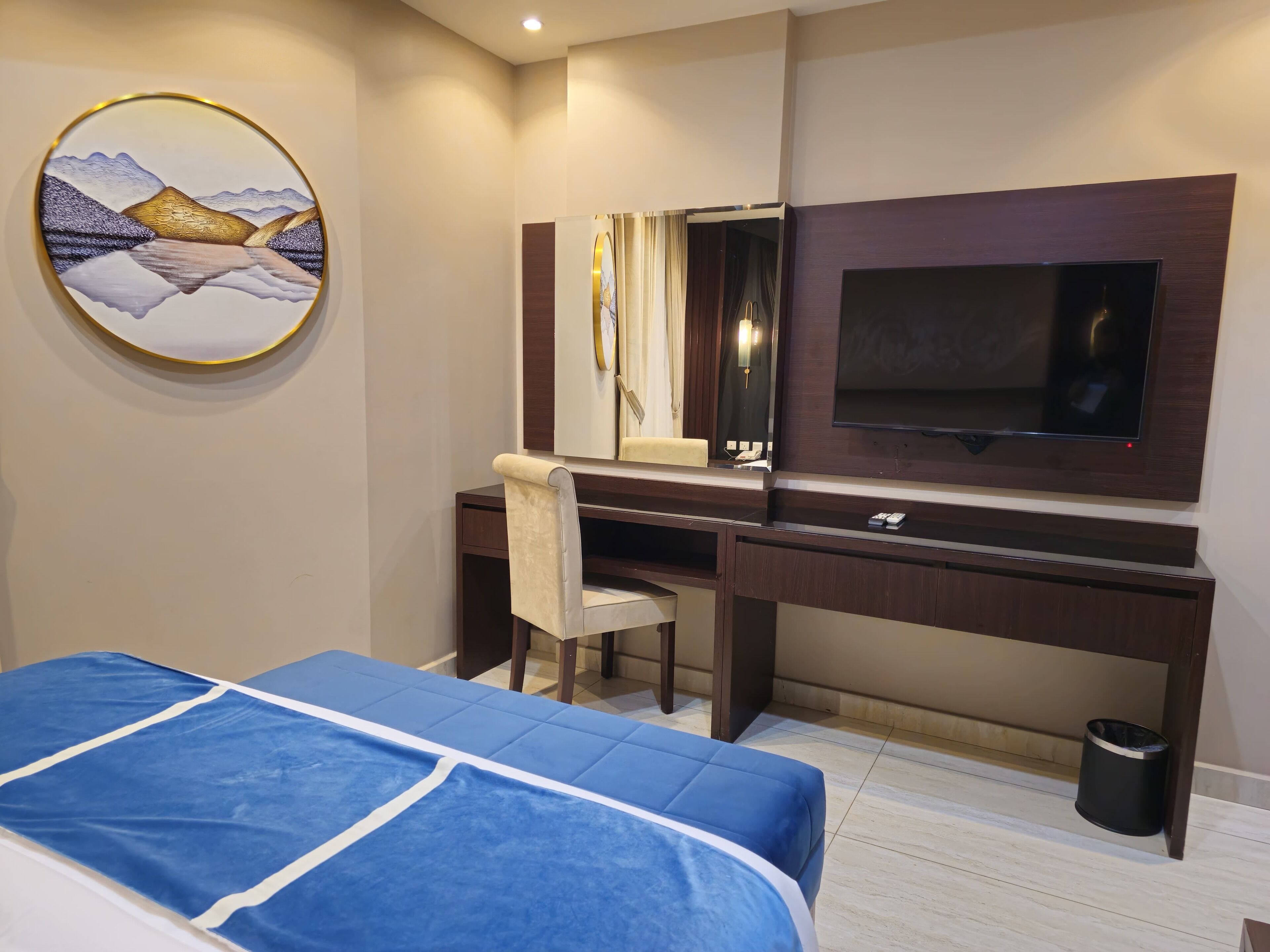 Quarto de hotel com cama, escrivaninha, televisão, espelho e uma obra de arte na parede.
