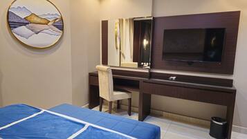 Quarto de hotel com cama, escrivaninha, televisão, espelho e uma obra de arte na parede.