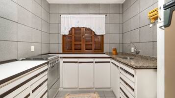 Cozinha compacta com armários brancos, fogão embutido, janela com cortina e bancada de granito.