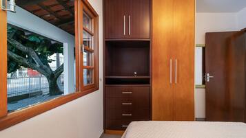 Quarto com cama, guarda-roupa de madeira e janela com vista para uma árvore e cerca.
