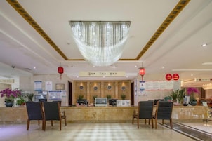Uma área de recepção de hotel com lustre, balcão de atendimento, cadeiras e cartazes informativos.