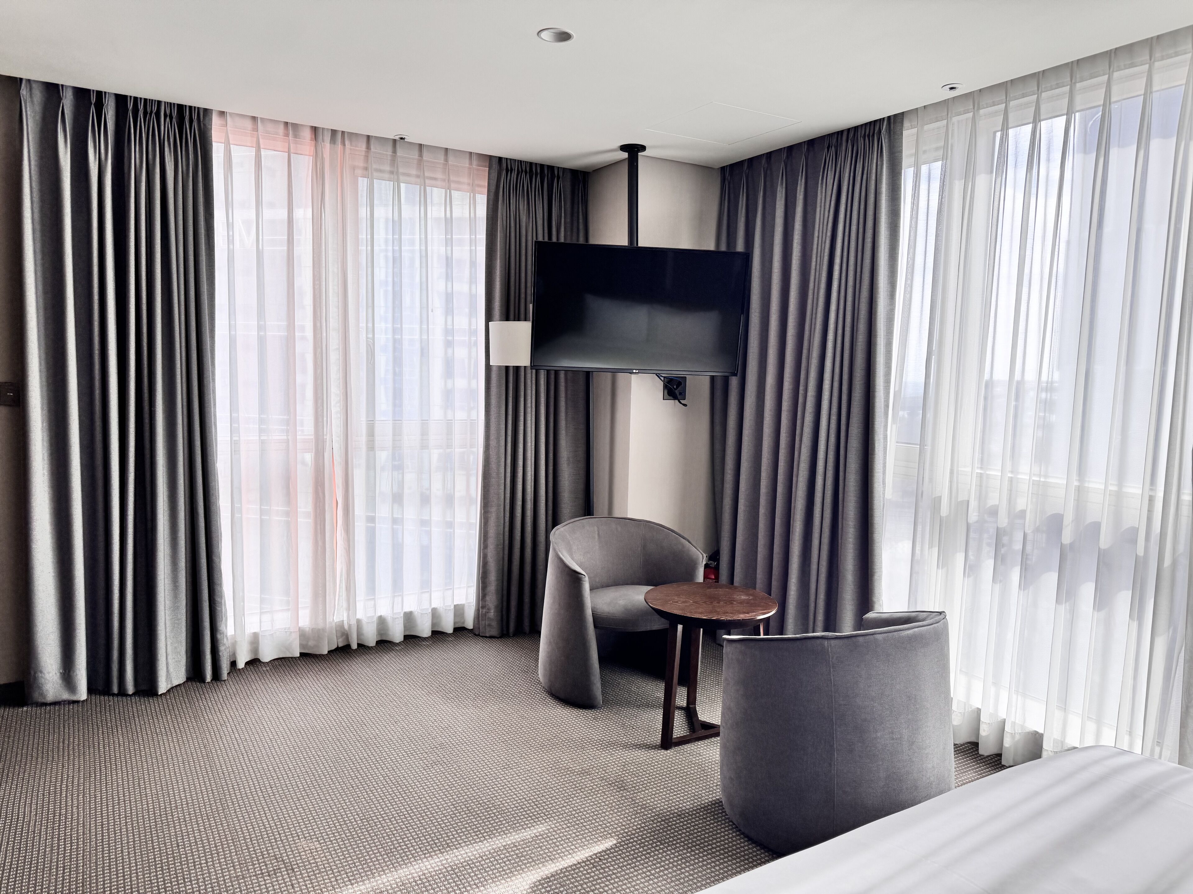 Quarto de hotel moderno com uma cama grande, área de estar com uma poltrona e uma mesa pequena, e uma TV de tela plana fixada na parede.