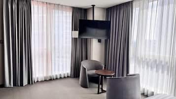 Quarto de hotel moderno com uma cama grande, área de estar com uma poltrona e uma mesa pequena, e uma TV de tela plana fixada na parede.