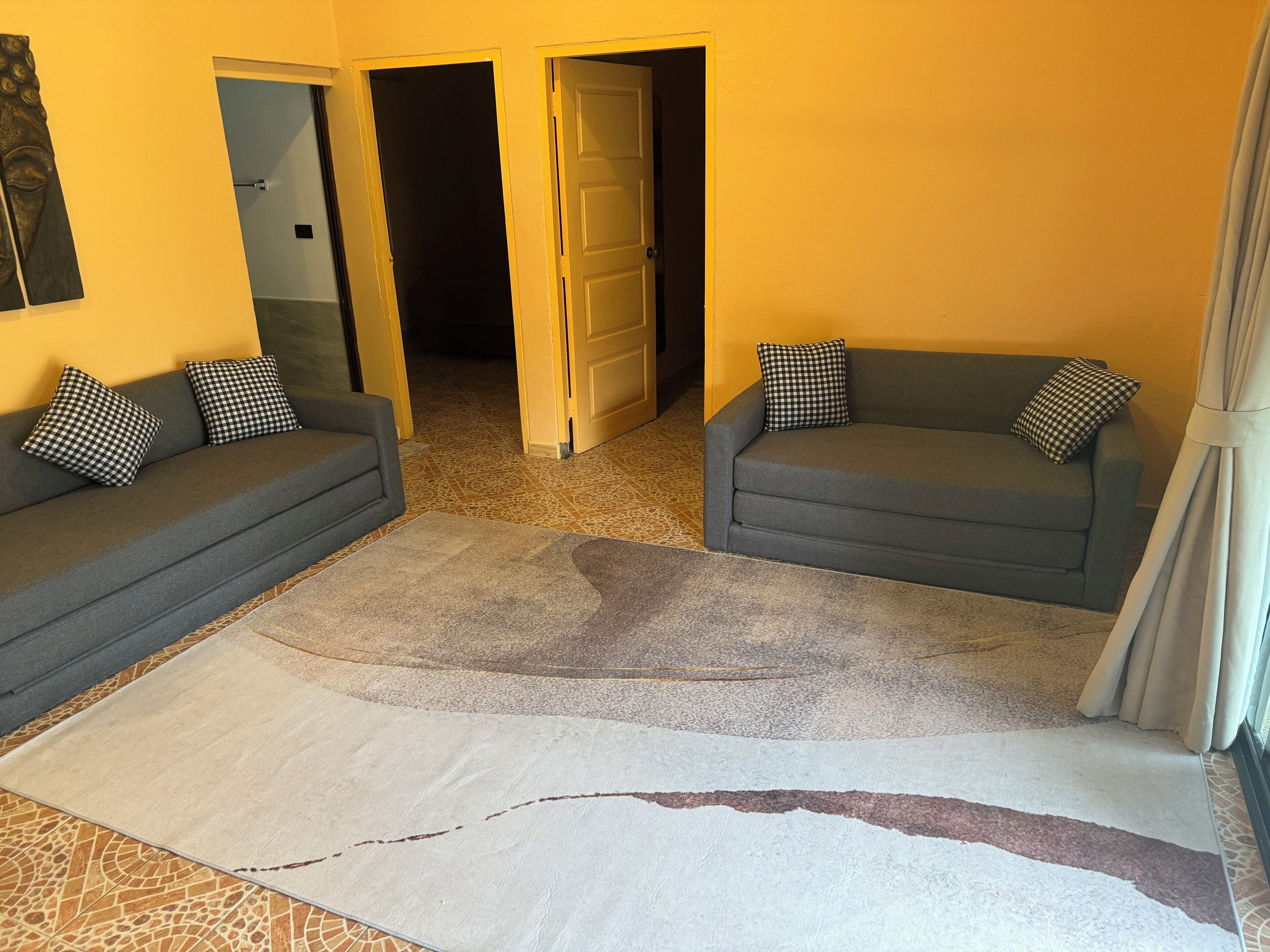 Sala de estar com dois sofás cinzas, almofadas xadrez, um tapete grande e uma porta que dá para outro cômodo.