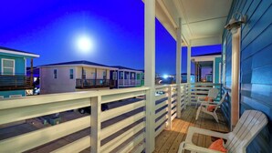 Varanda com grades brancas, piso de madeira e vista para uma fileira de casas sob um céu noturno com a lua visível.