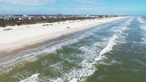 Uma praia com uma extensa faixa de areia, ondas e uma fileira de edifícios ao fundo.