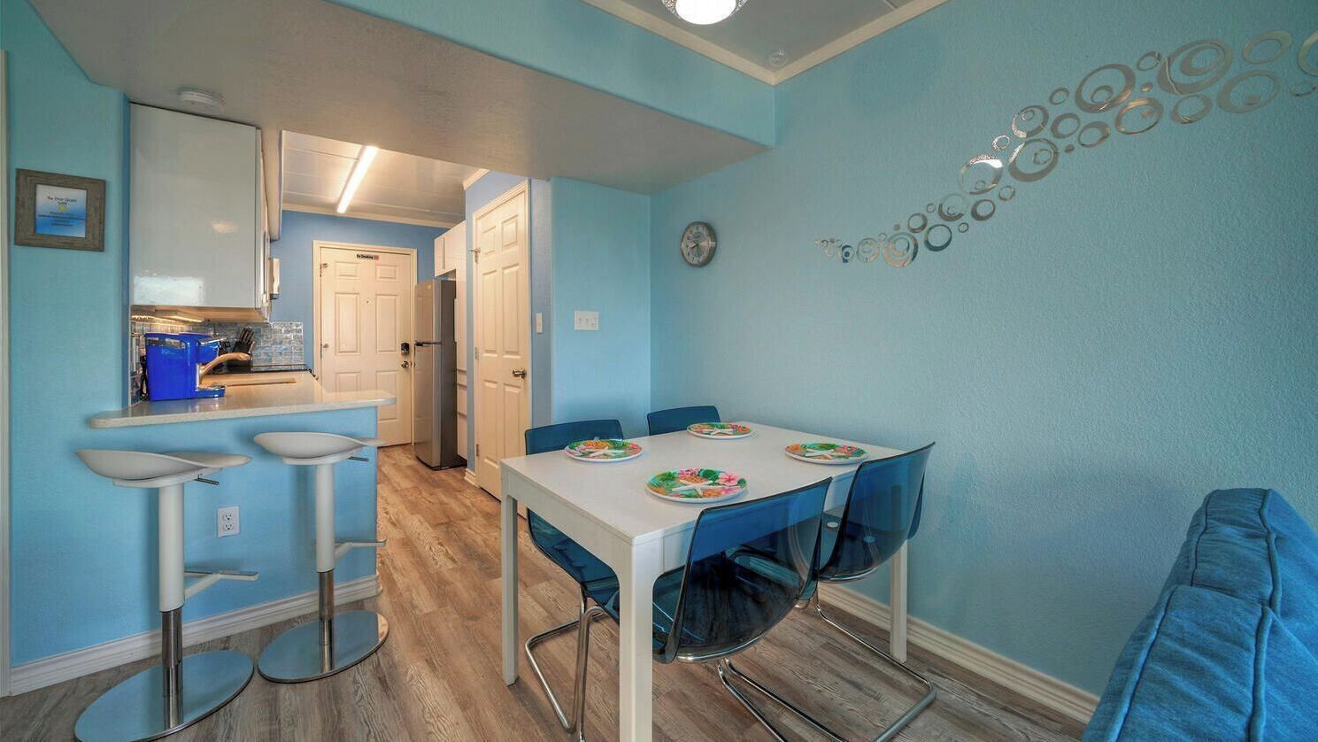 Uma área de refeições com uma mesa branca e cadeiras azuis, cozinha ao fundo e uma parede azul com um desenho circular.