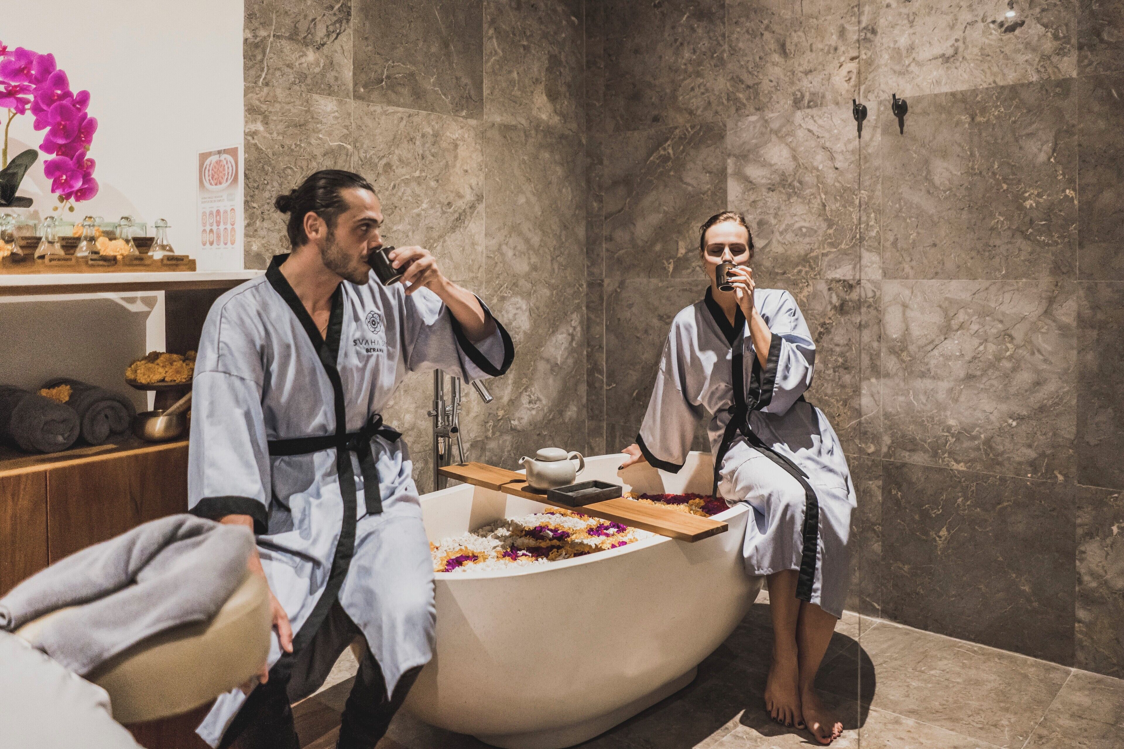 Ambiente de spa com banheira, toalhas e uma estante decorativa com flores.